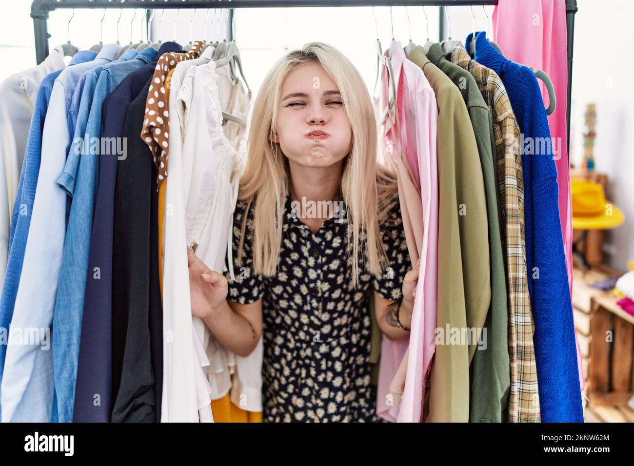 Young blonde woman searching clothes on clothing rack puffing cheeks ...