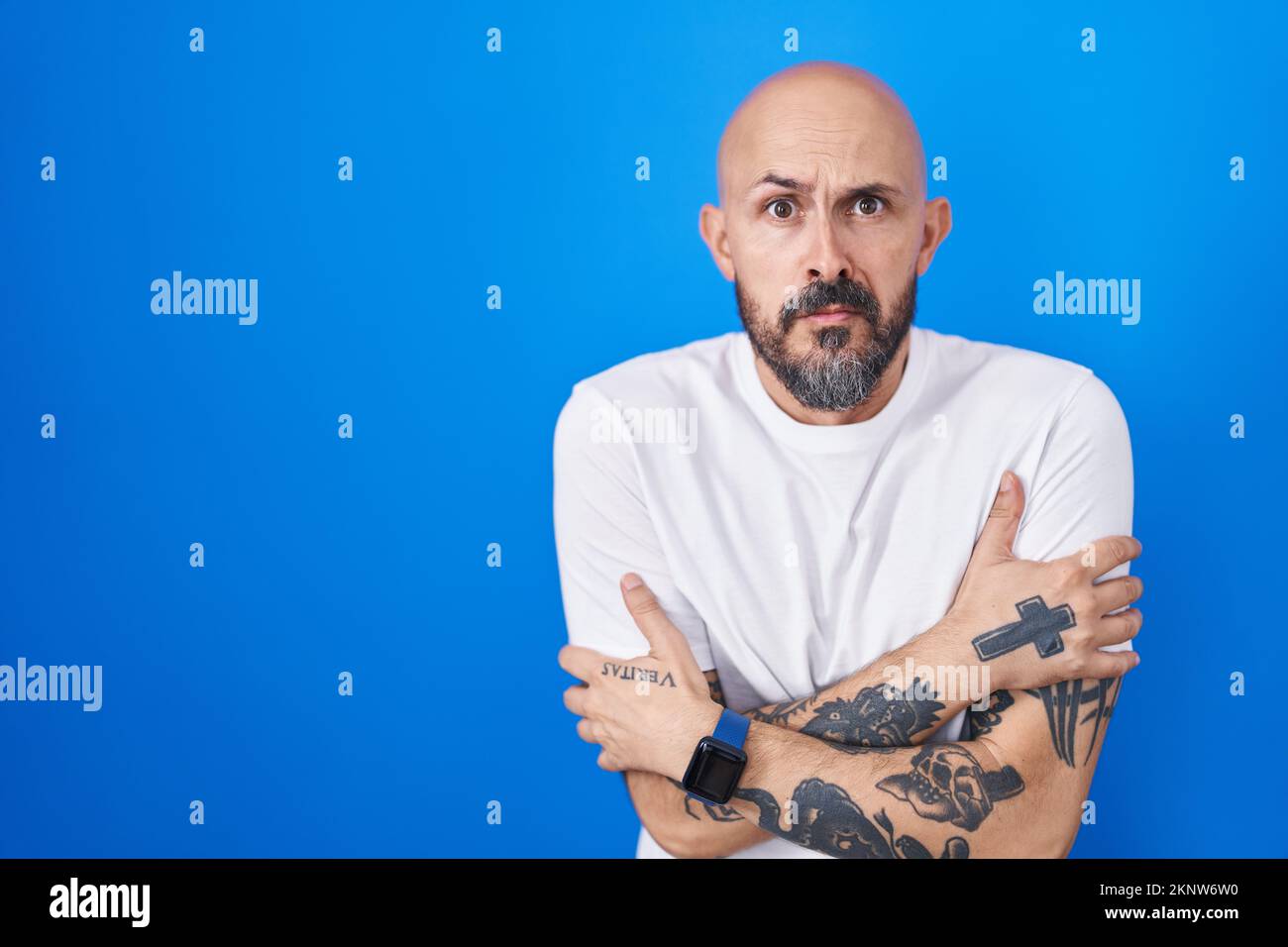 Hispanic man with tattoos standing over blue background shaking and ...