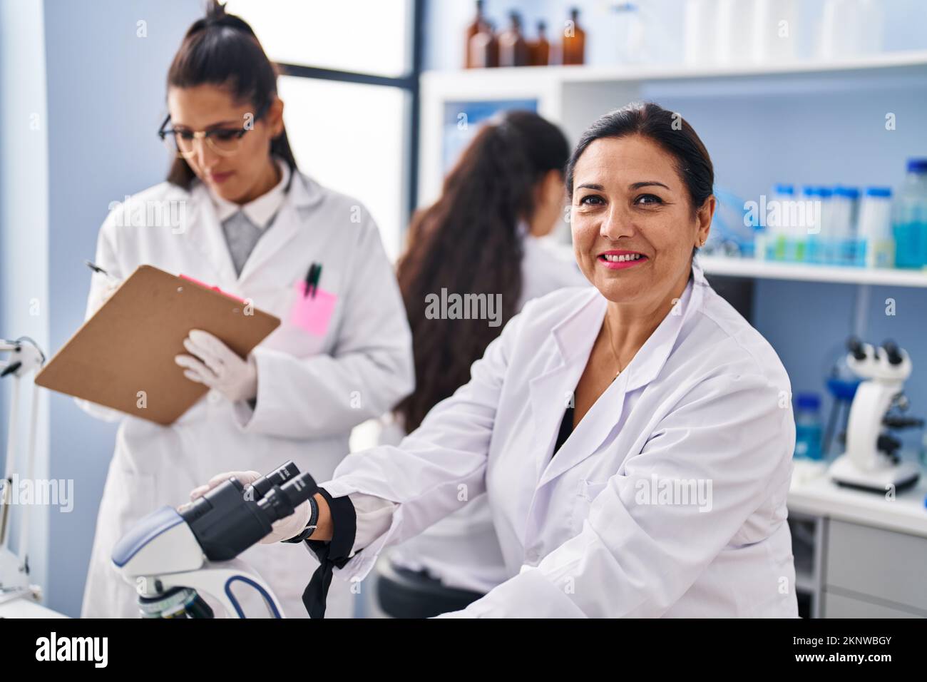 Three woman scientists using microscope write on checklist at ...