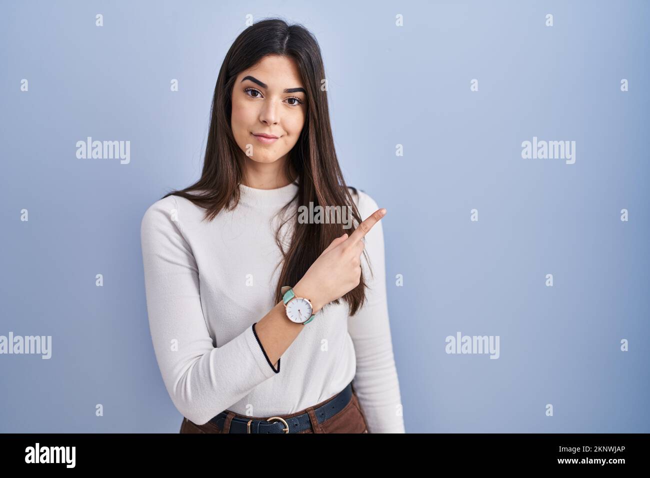 Young brunette woman standing over blue background pointing with hand ...