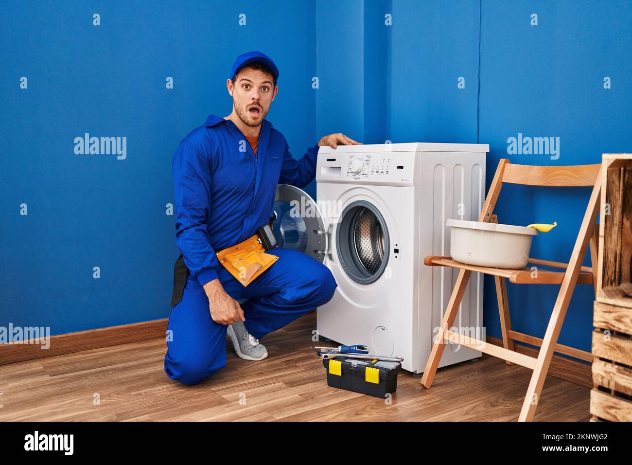 Young hispanic man working on washing machine scared and amazed with ...