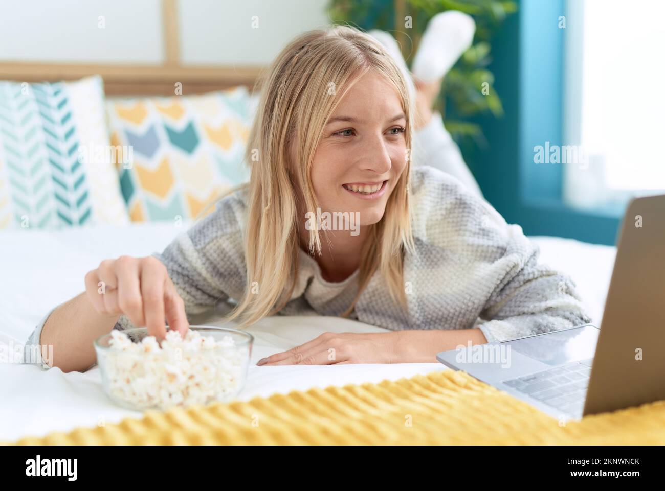 Young blonde woman watching movie on laptop lying on bed at bedroom ...