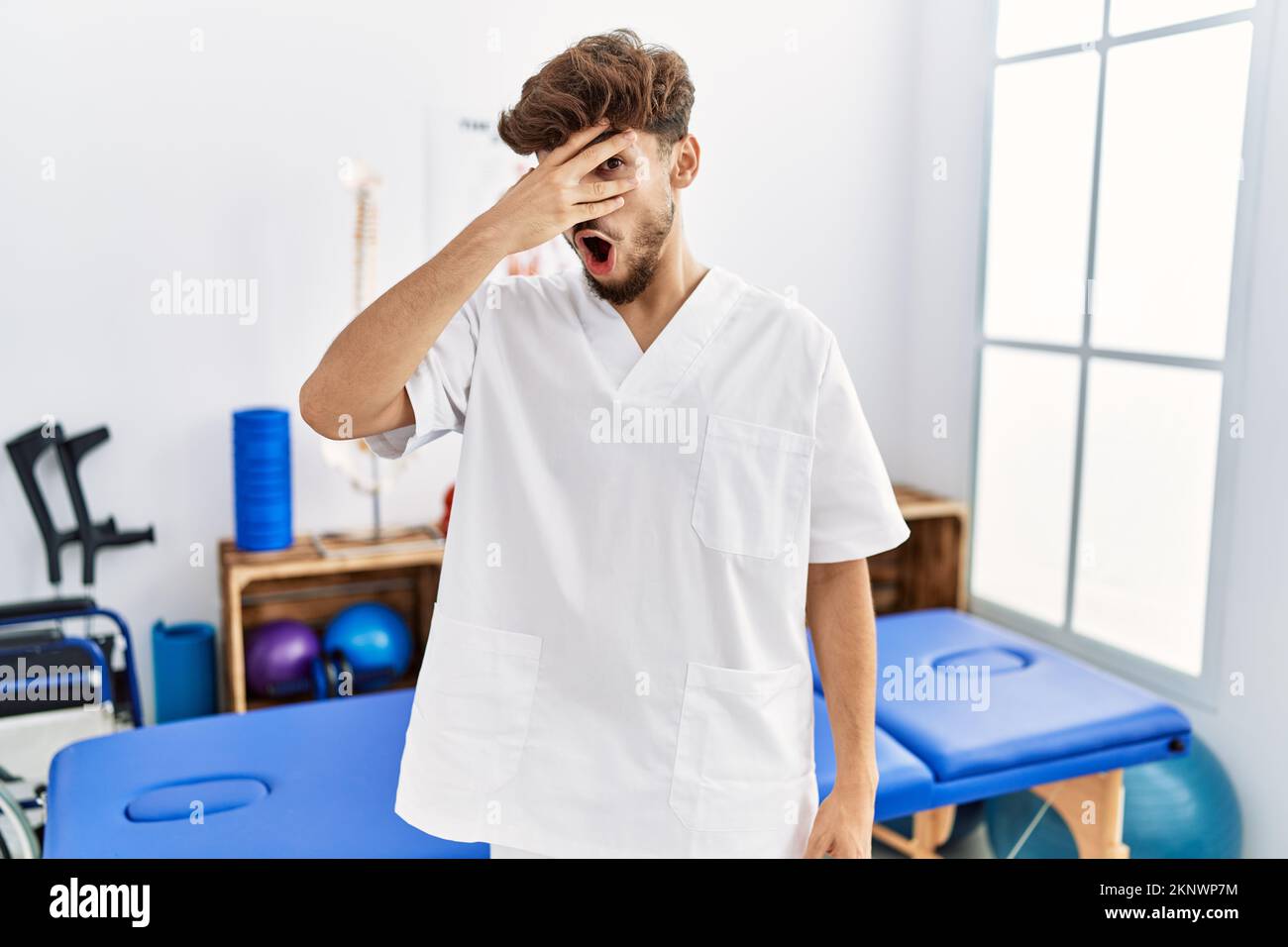 Young arab man working at pain recovery clinic peeking in shock ...