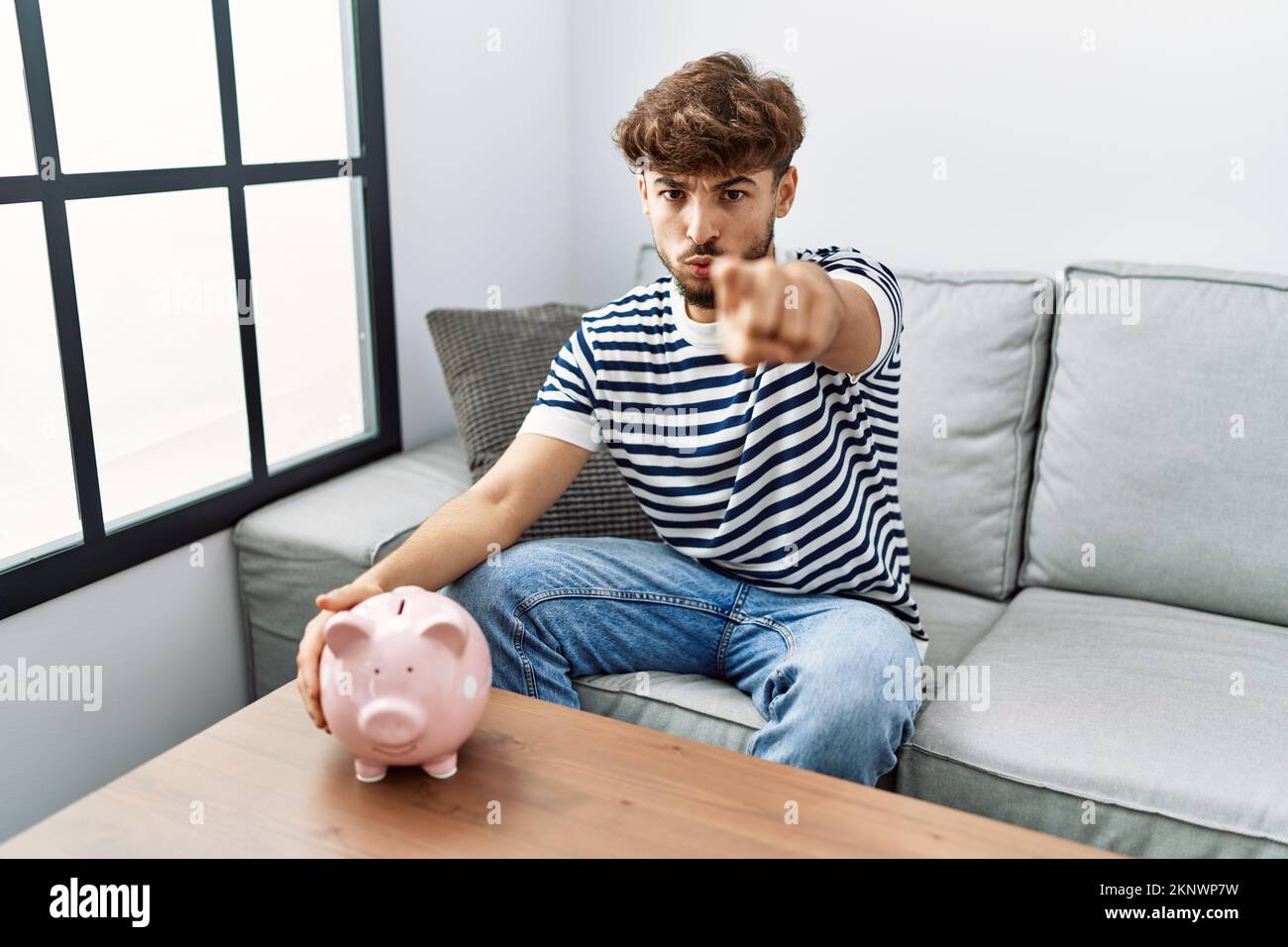 Young arab man holding piggy bank pointing with finger to the camera ...