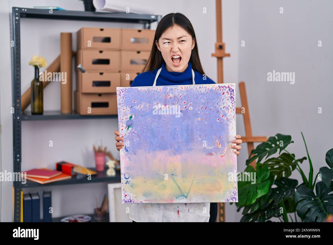 Chinese young woman holding canvas at art studio angry and mad ...