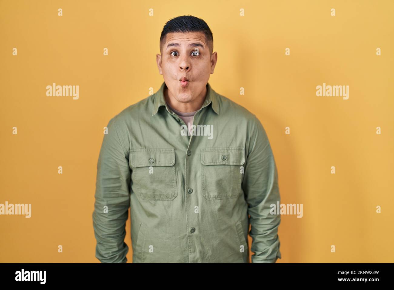 Hispanic young man standing over yellow background making fish face ...