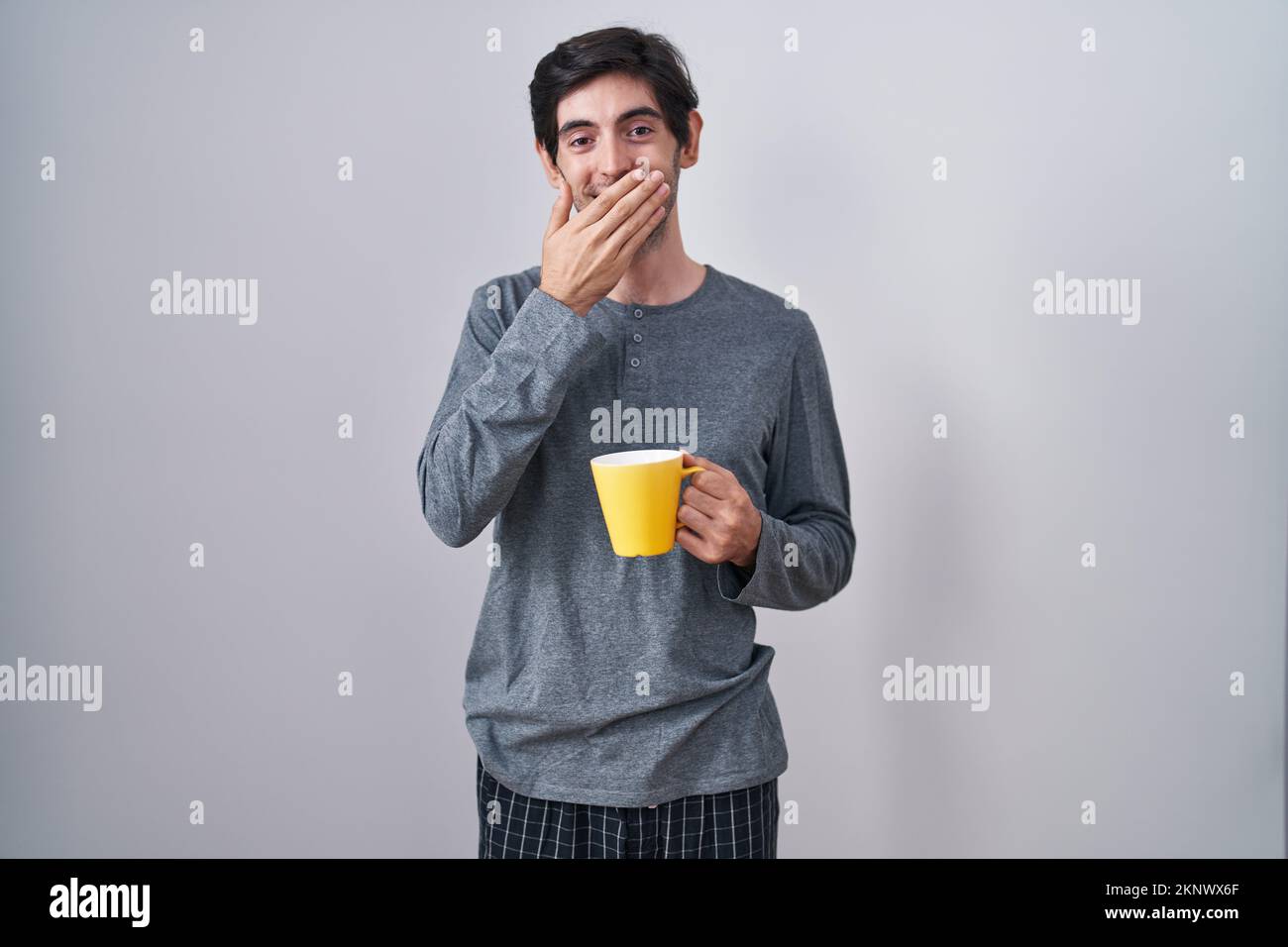 Young hispanic man wearing pajama drinking a cup of coffee laughing and ...