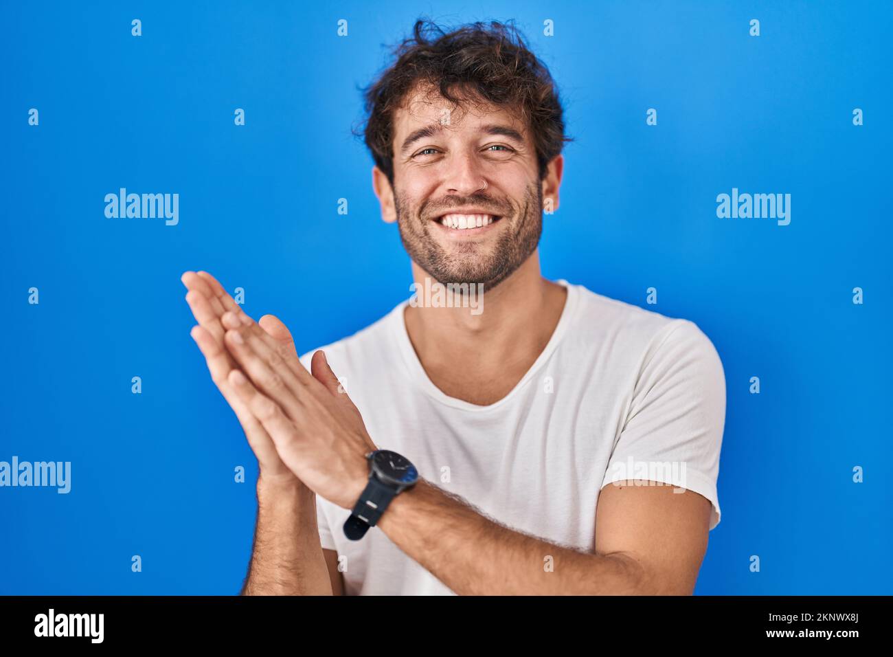 Hispanic young man standing over blue background clapping and ...