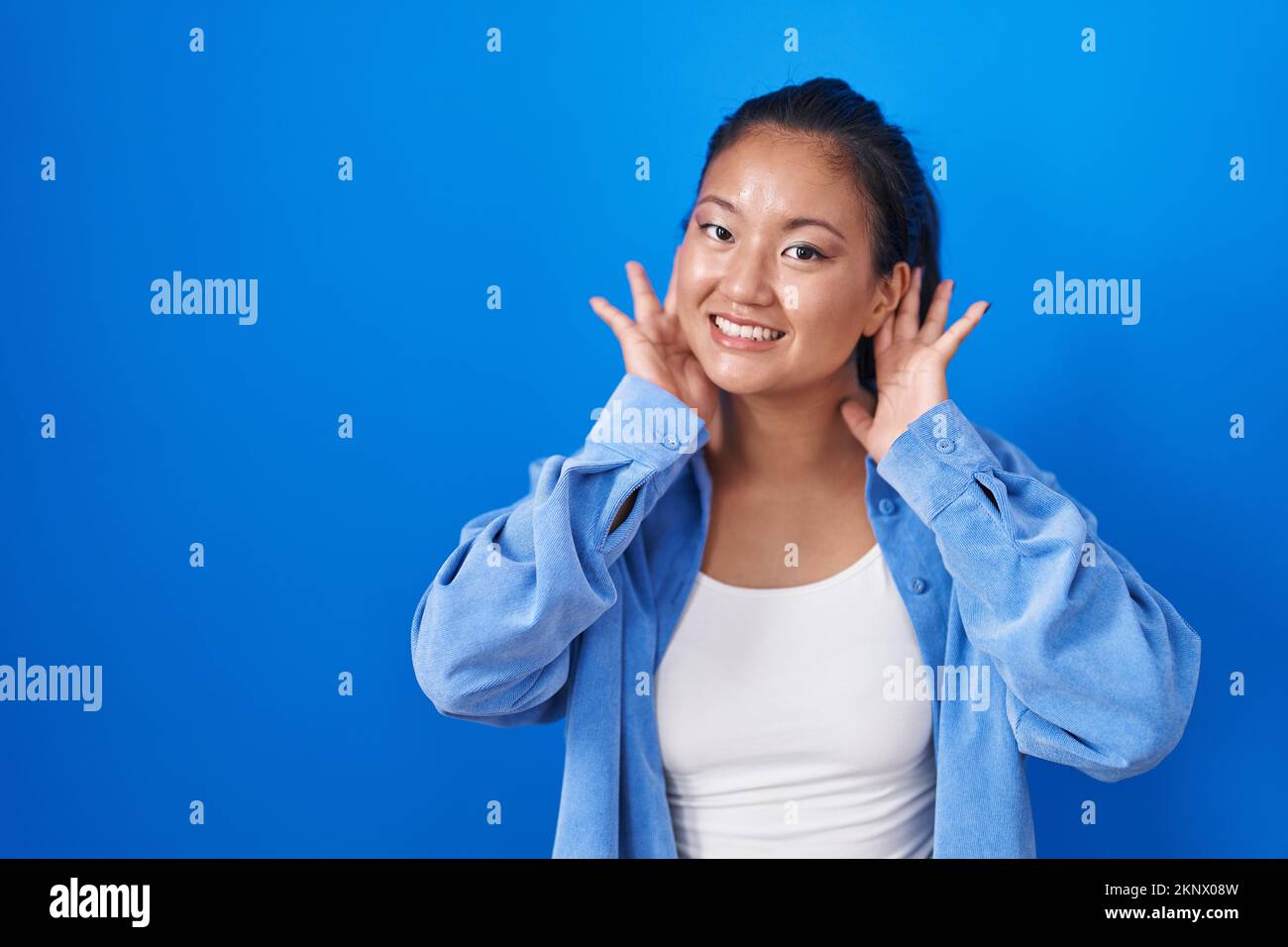 Asian young woman standing over blue background smiling pulling ears ...