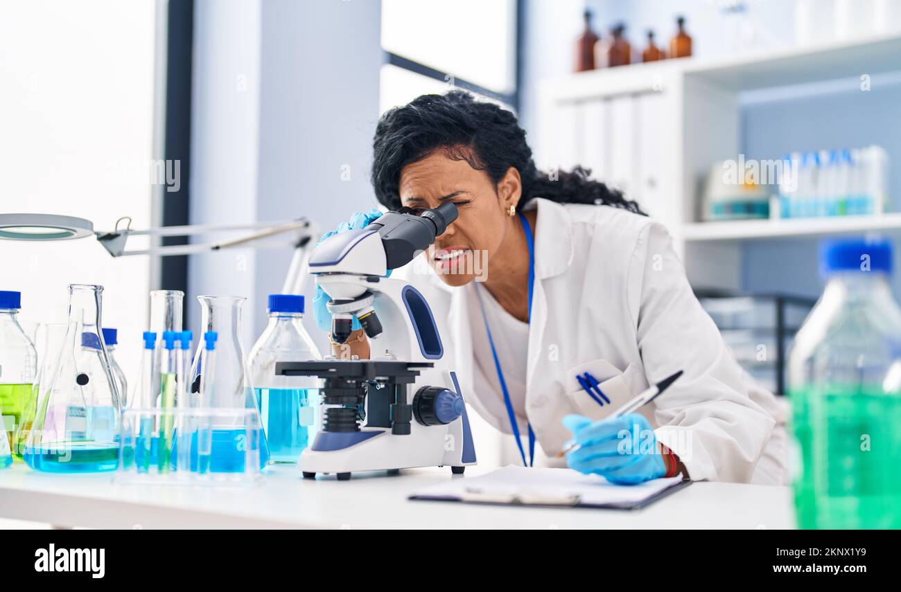 African american woman wearing scientist uniform using microscope write on document at ...