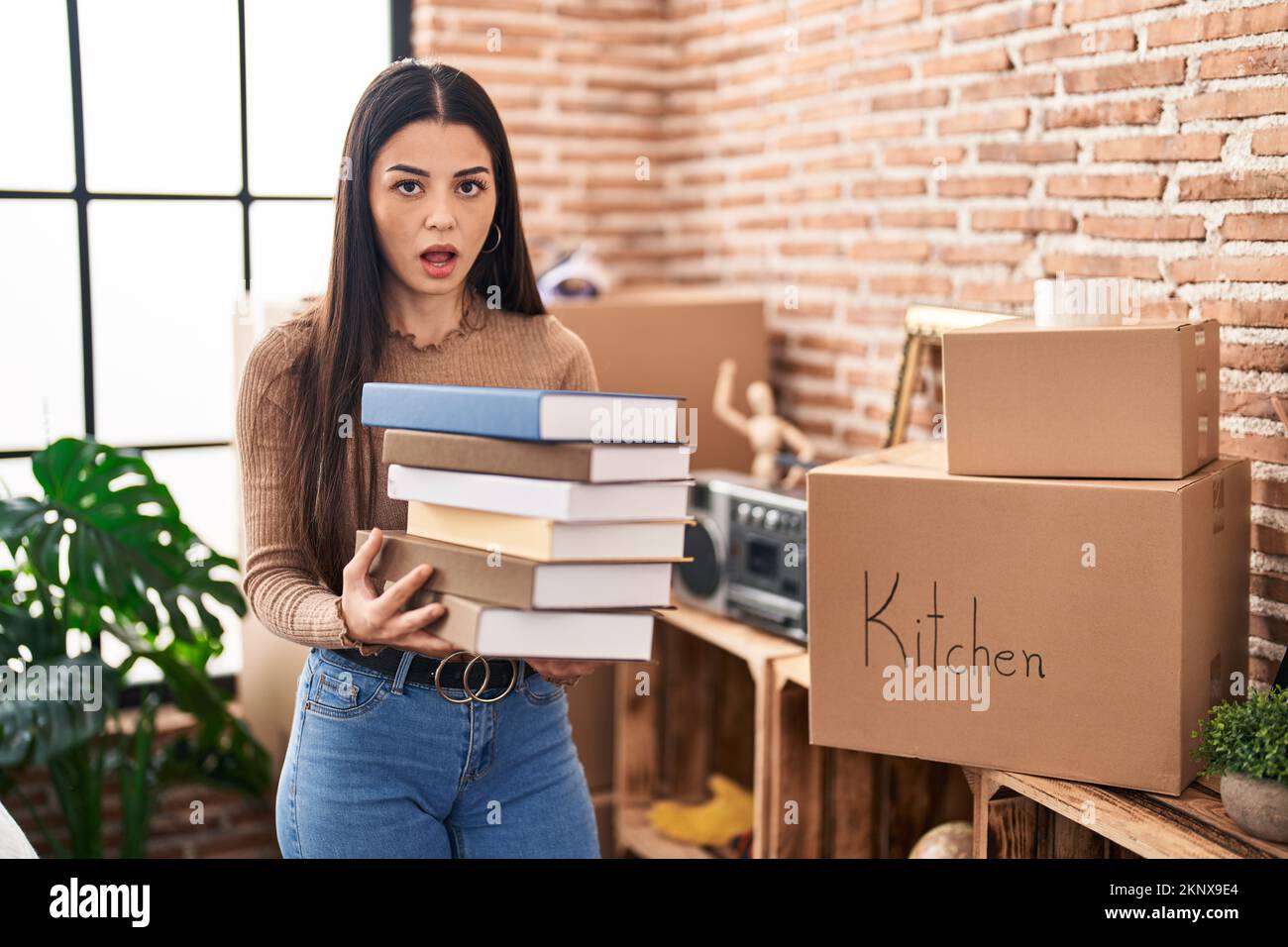 Young woman holding books at home in shock face, looking skeptical and ...