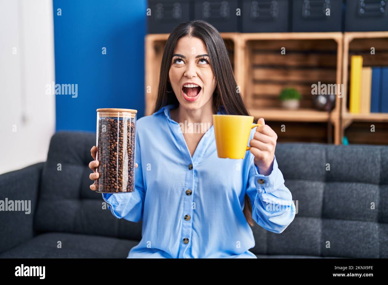 Young brunette woman holding coffee beans and cup of coffee angry and ...