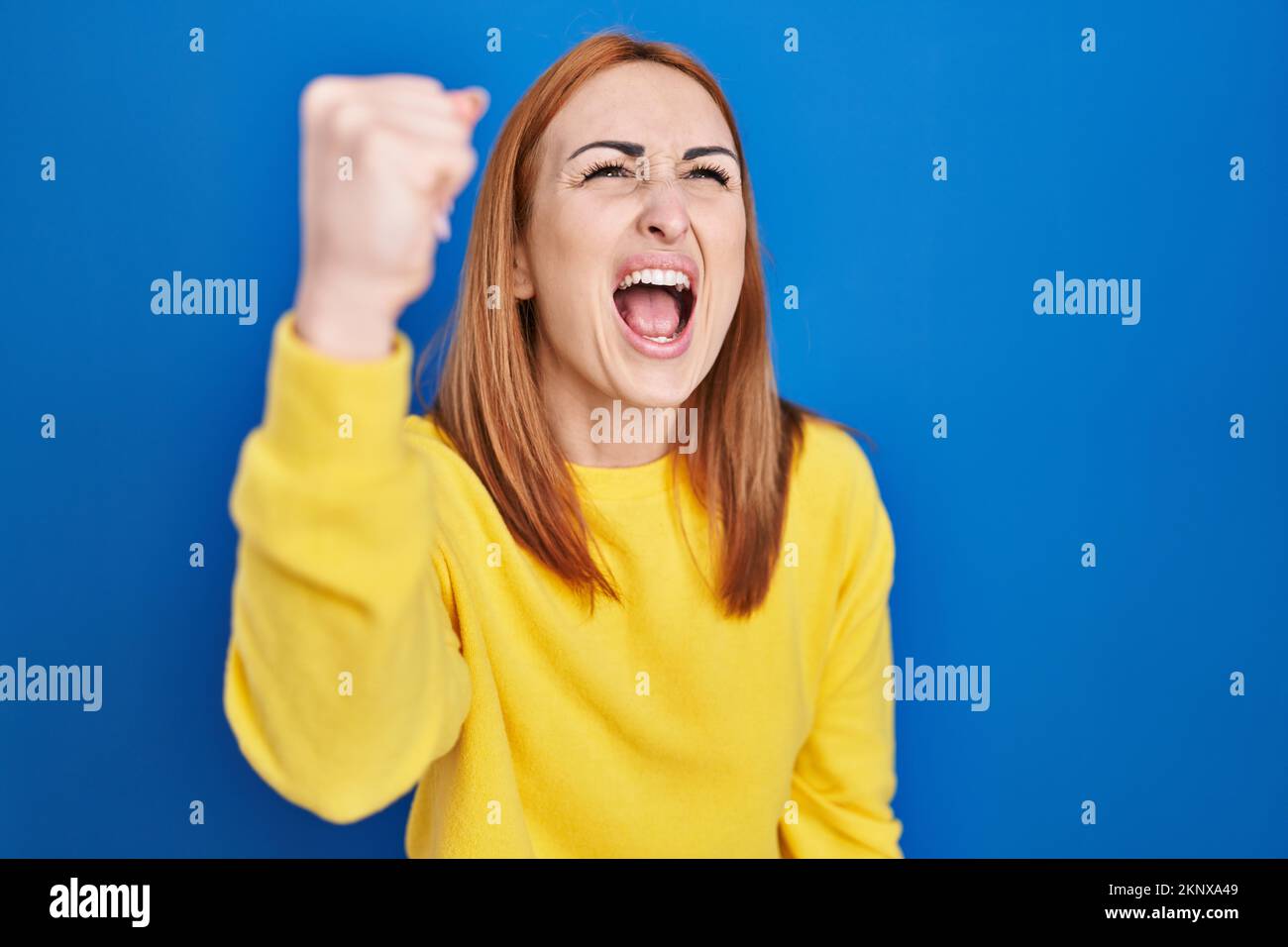Young woman standing over blue background angry and mad raising fist ...