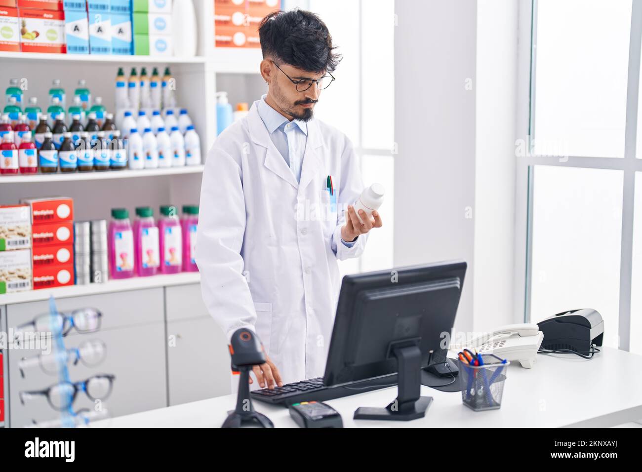 Young hispanic man pharmacist holding pills bottle using computer at ...