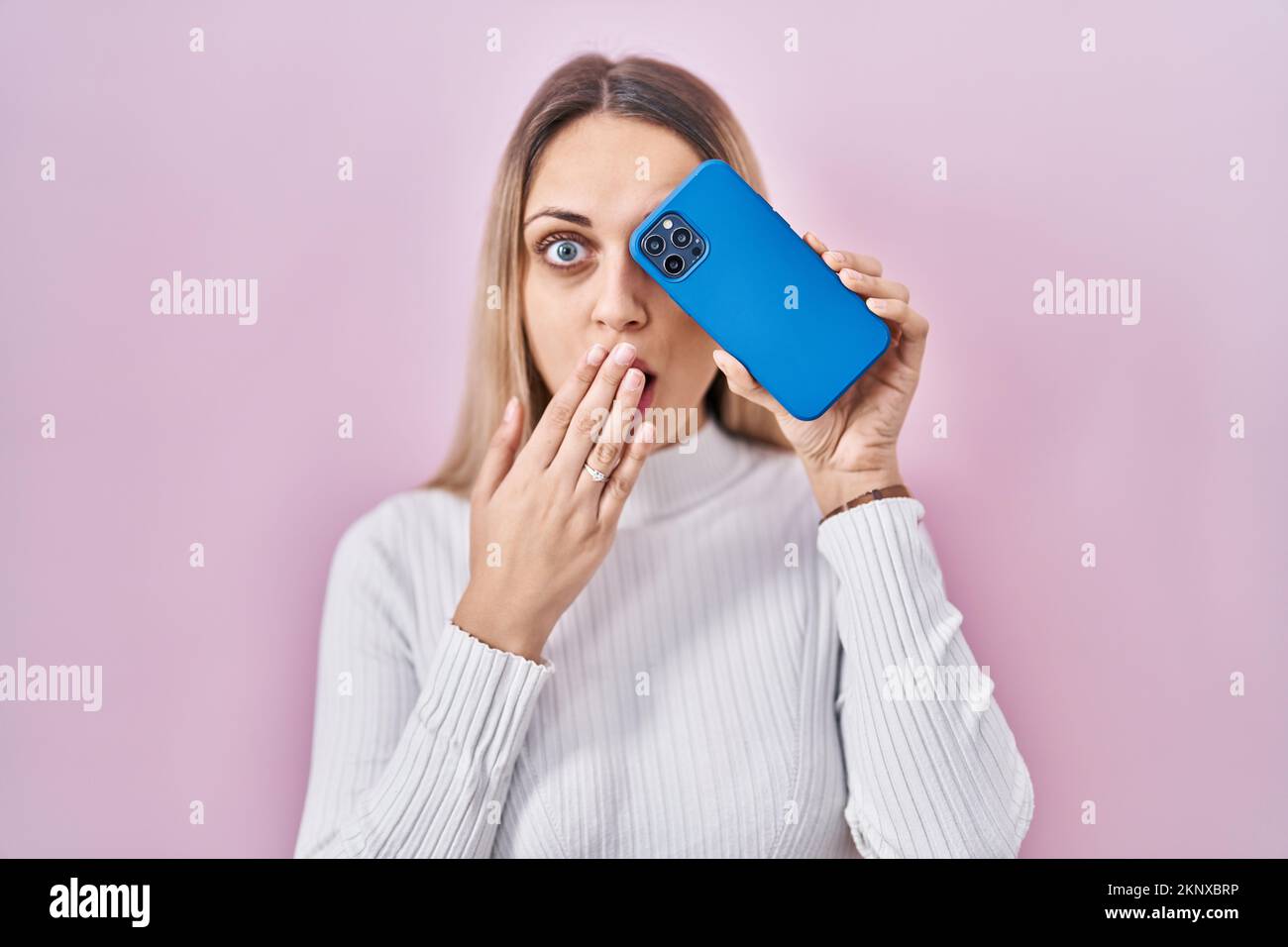 Young blonde woman holding smartphone covering eye covering mouth with ...