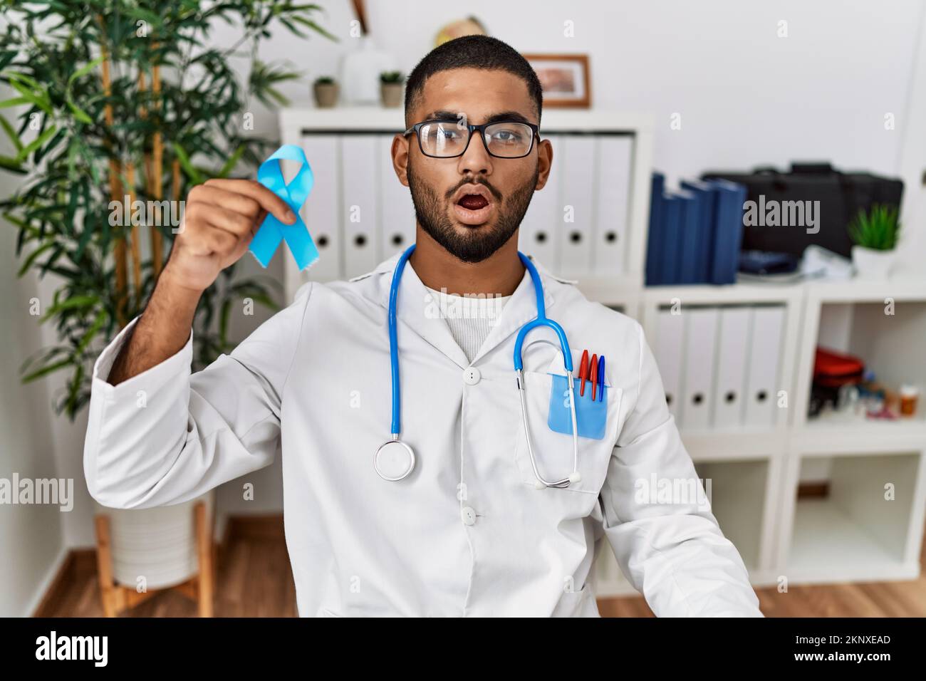 Young indian doctor holding blue ribbon scared and amazed with open ...
