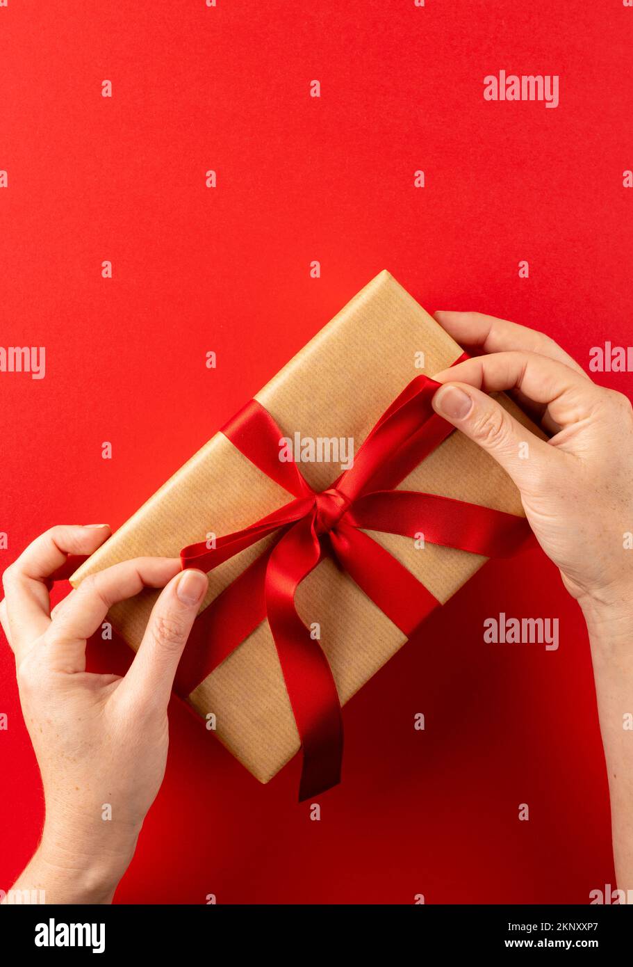 Vertical image of hands tying gift in brown paper with red ribbon, on ...