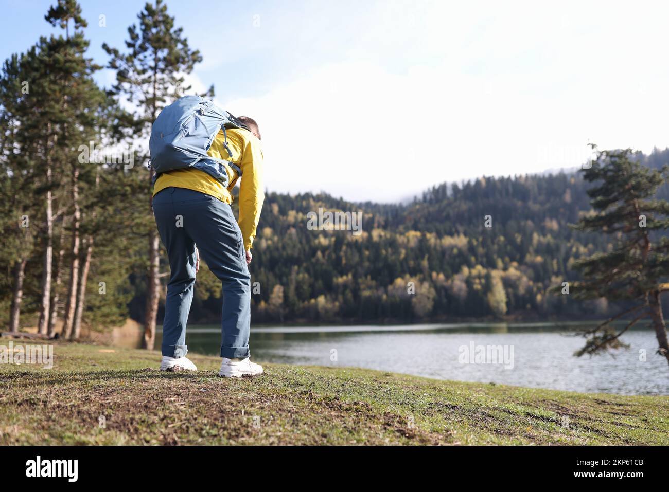 Tired male traveler with heavy backpack bending over near forest lake ...