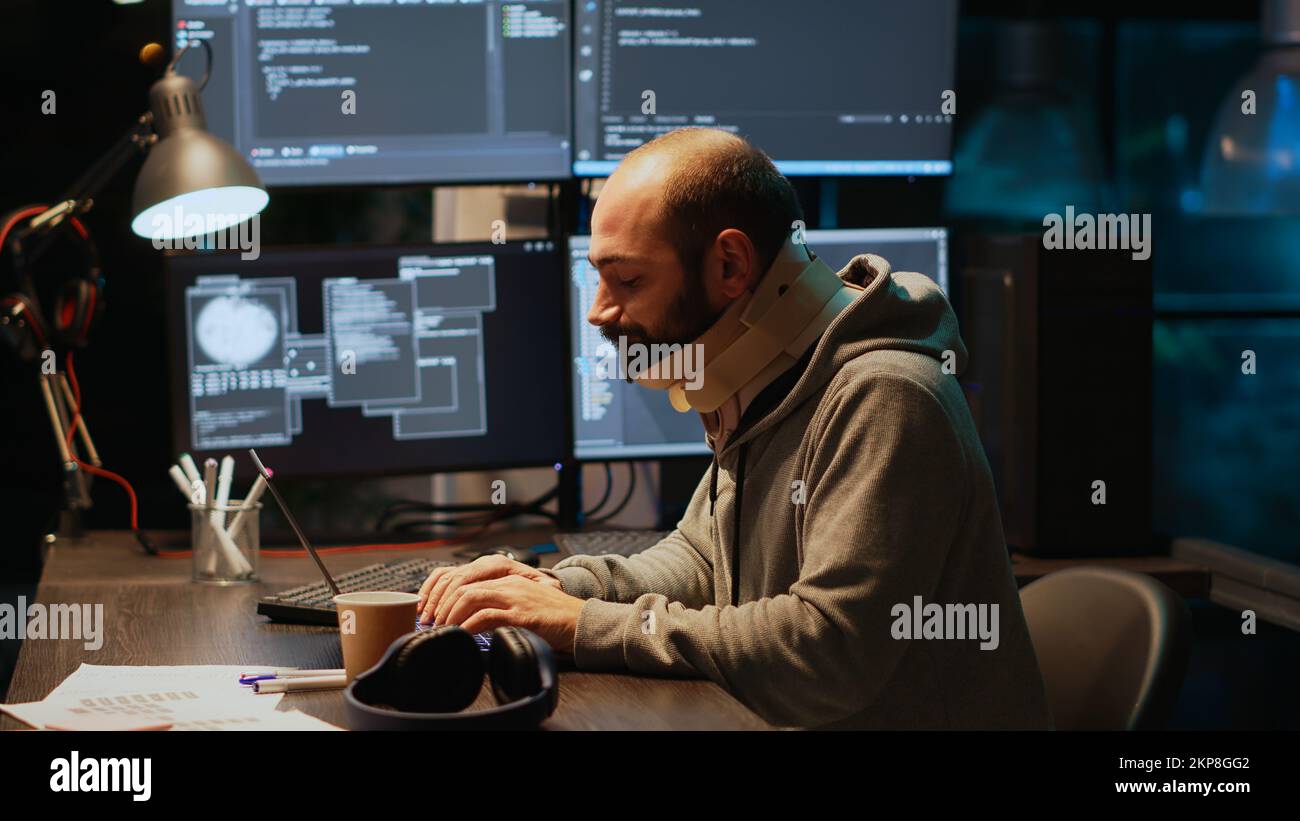 Adult programmer with neck collar foam coding html script, using laptop and multiple monitors at ...