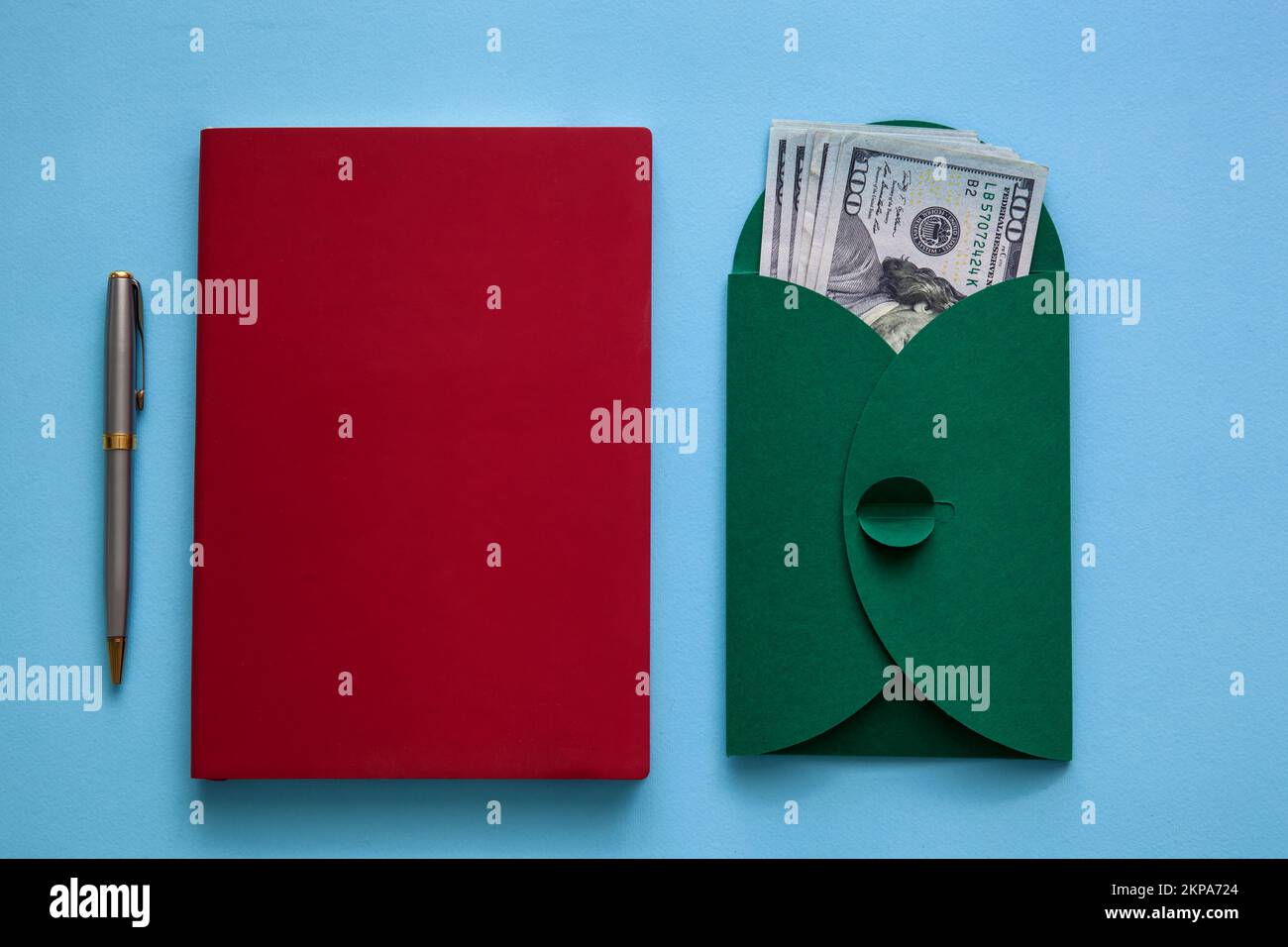 One hundred dollar bills in a green envelope, a red diary and a pen on ...