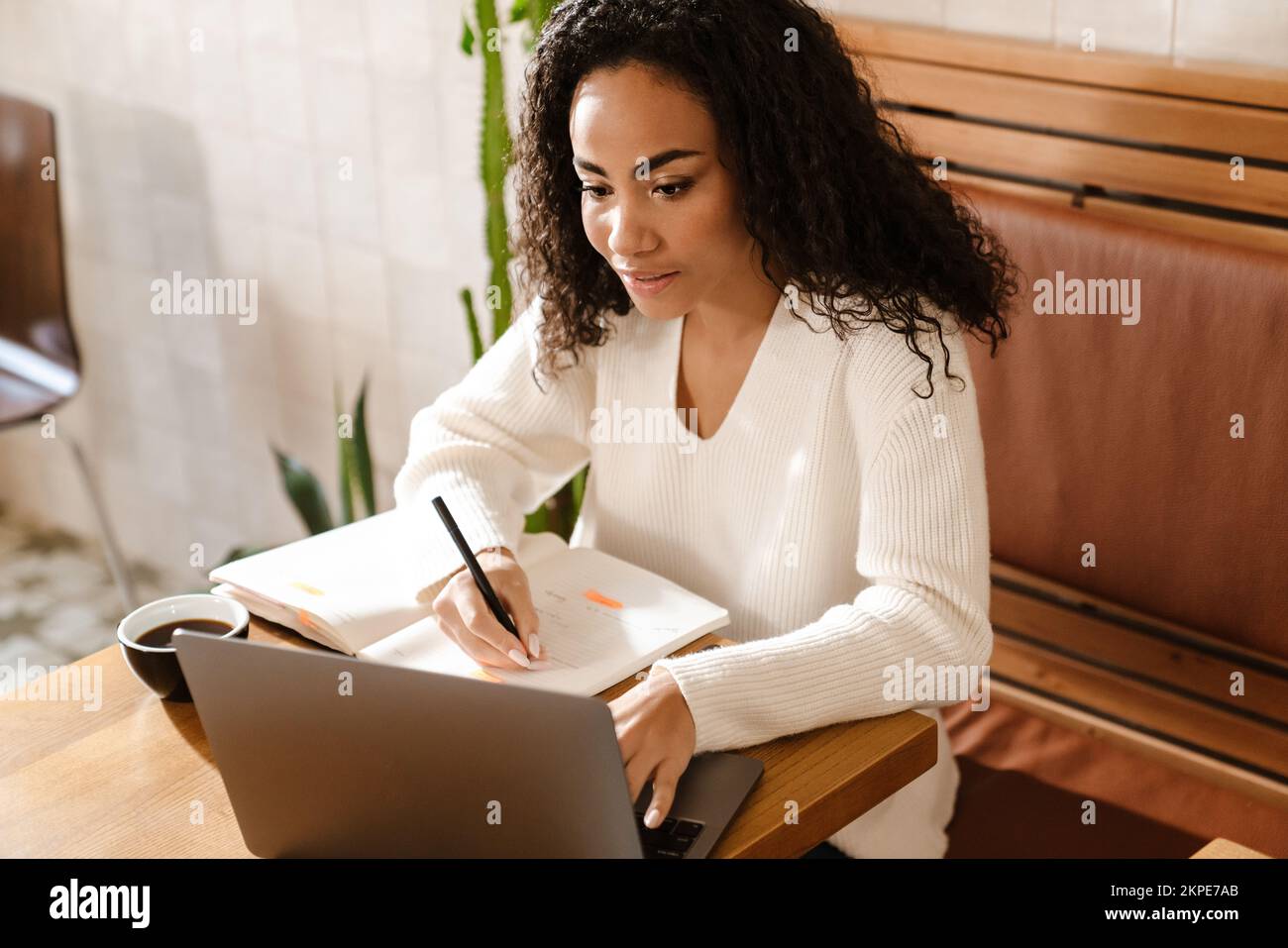 Young black woman writing down notes while working with laptop in cafe ...