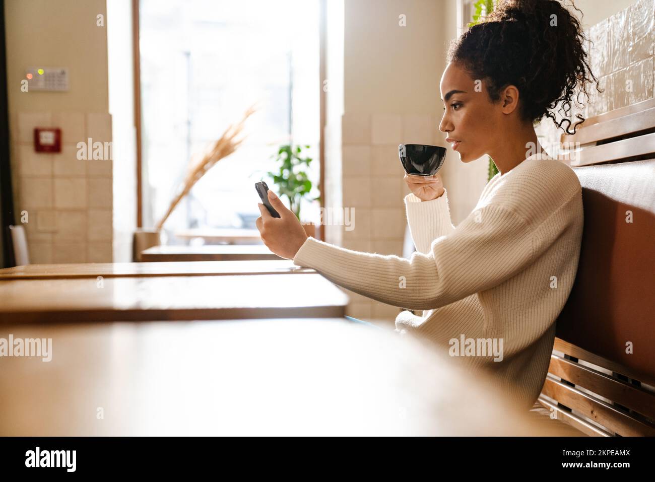Young black woman using mobile phone while drinking coffee in cafe ...
