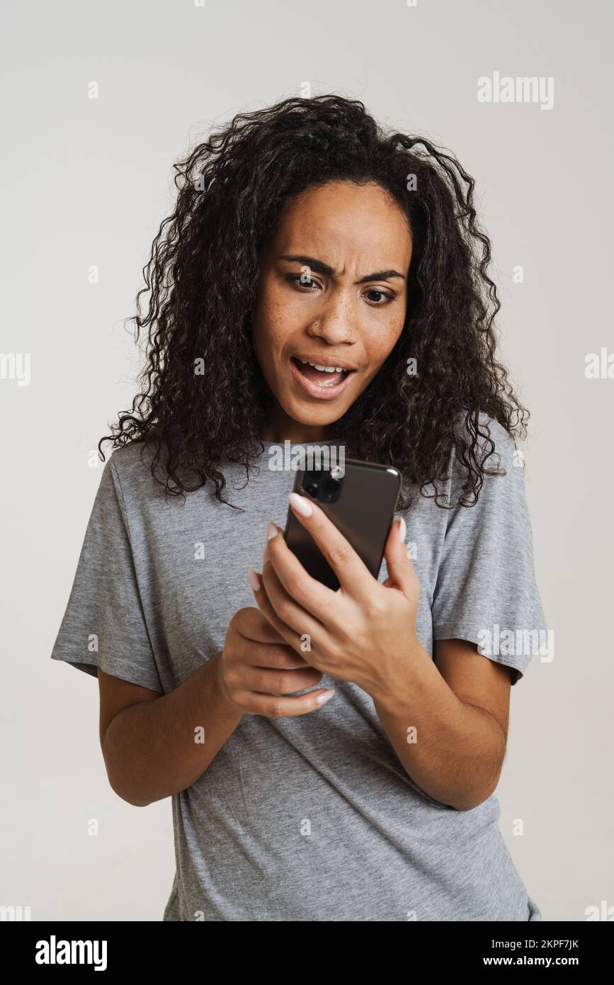 Young black woman using mobile phone and expressing surprise isolated ...