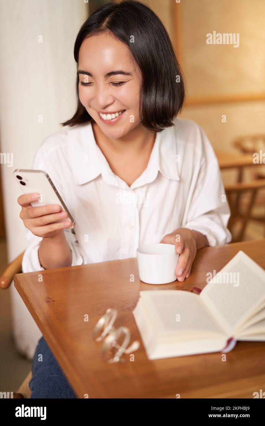 Happy asian woman sits in cafe with cup of coffee and book, answer ...