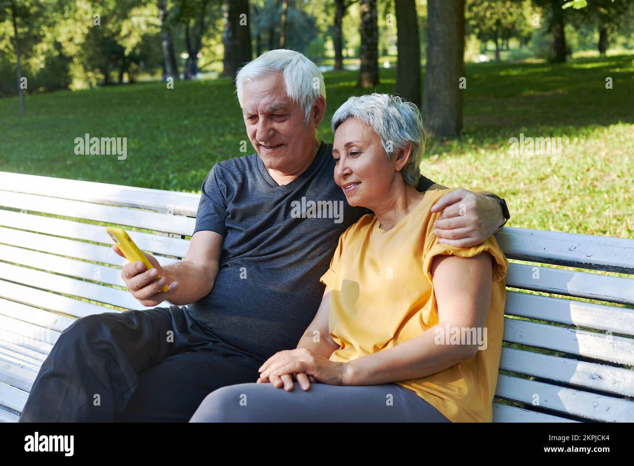 Mature spouses sit on bench in summer park with smart phone. Grey ...