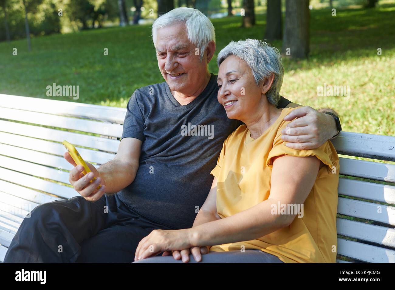 Mature spouses sit on bench in summer park with smart phone. Grey ...