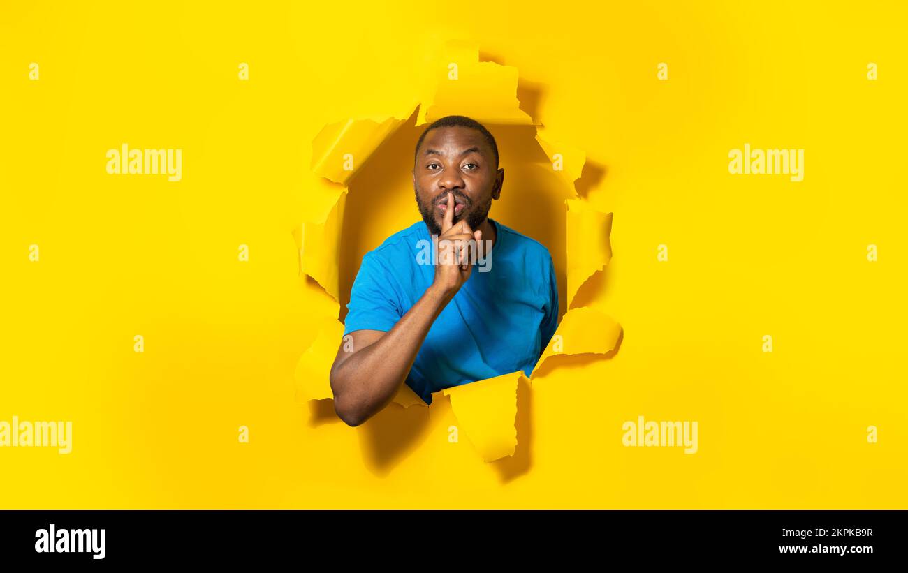Black man making silence gesture telling secret, breaking through hole ...