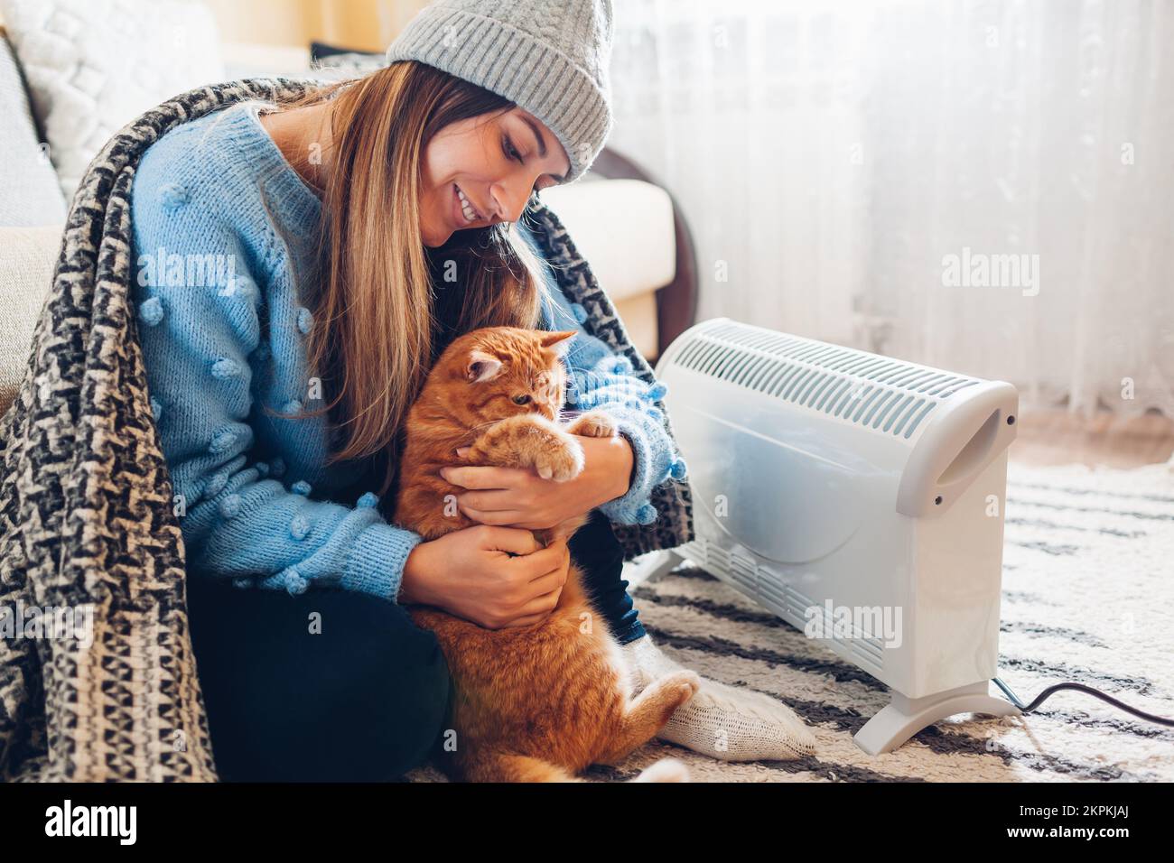 Using heater at home in cold winter. Woman playing with cat by electric ...