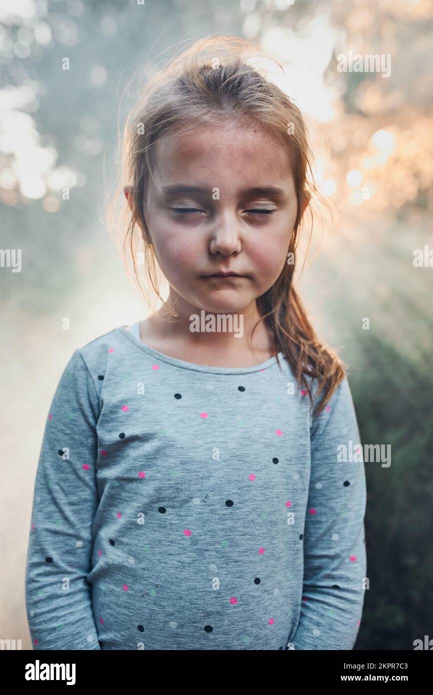 Portrait of sad little girl with closed eyes standing in smoke back to ...