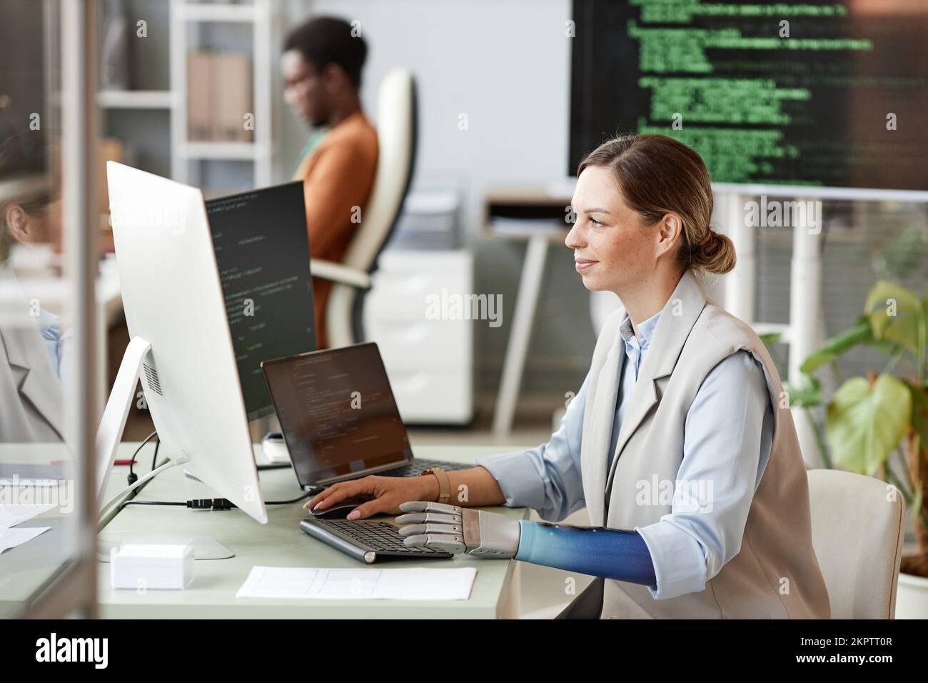 Smiling woman with bionic arm coding on computer when working on big project at research center ...