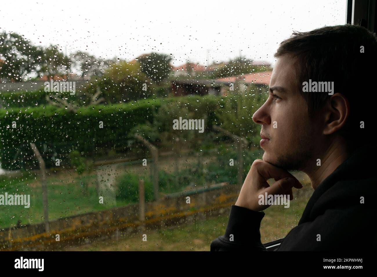 Profile portrait of young man looking out of a window on a rainy day ...