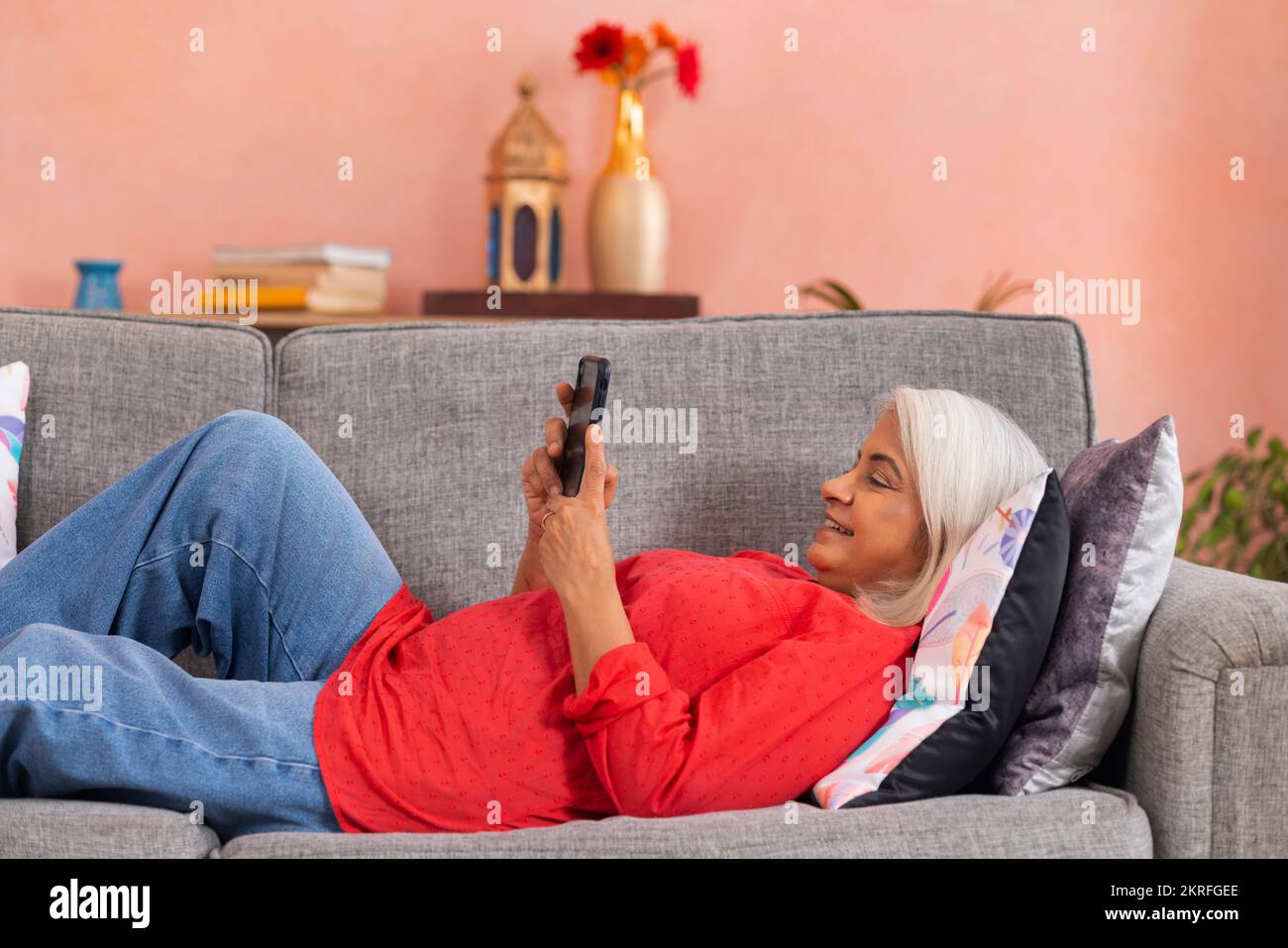 Old woman using mobile phone while lying down on sofa in living room ...