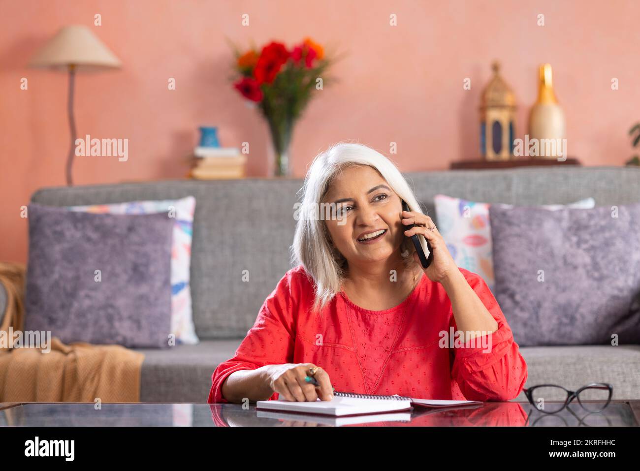 Old woman talking on mobile phone while writing on notepad at home ...