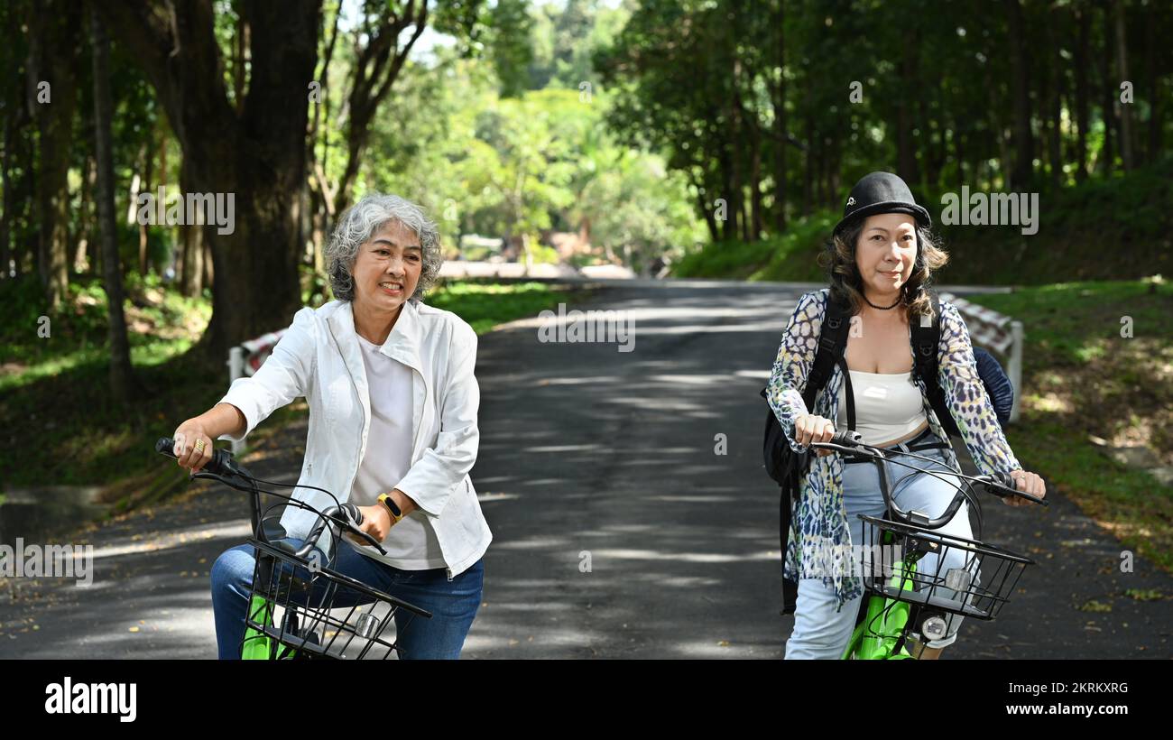 Two smiling middle aged women cycling in nature park on beautiful day ...