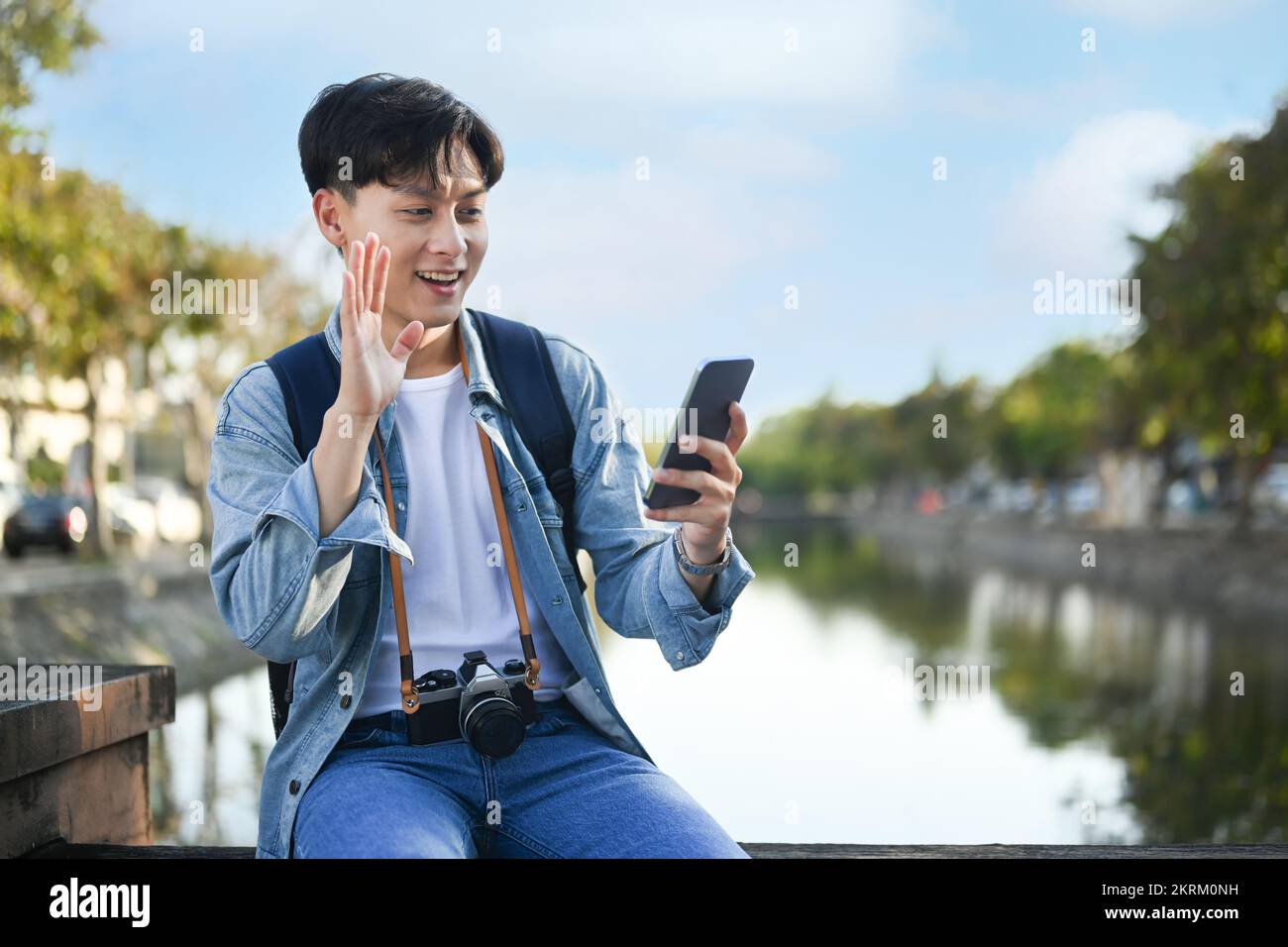 Young asian male blogger sitting on bridge bench near river and holding smartphone recording ...