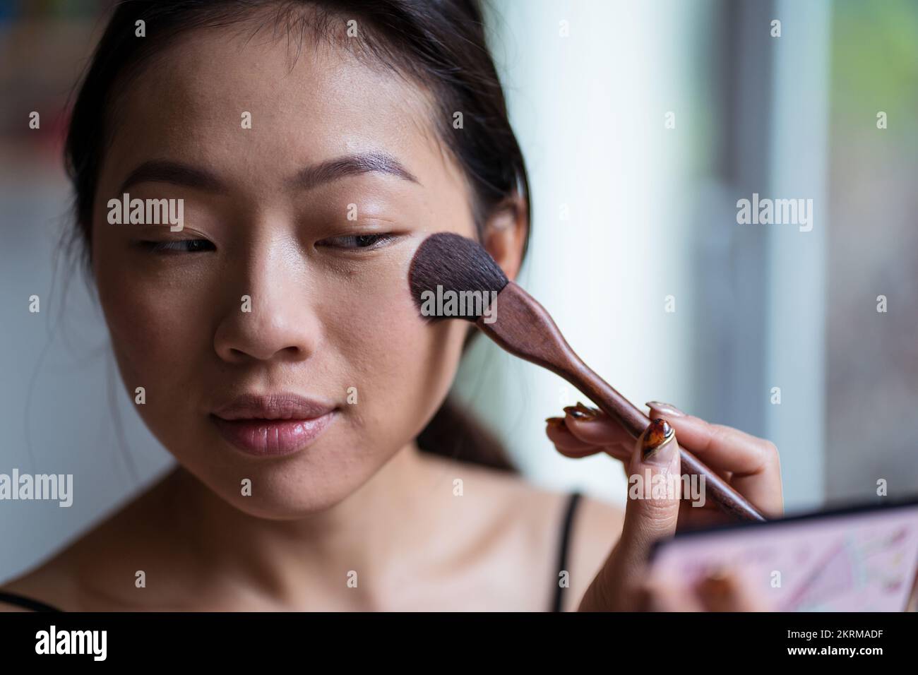 Crop ethnic female spreading powder and blush on cheeks while looking ...
