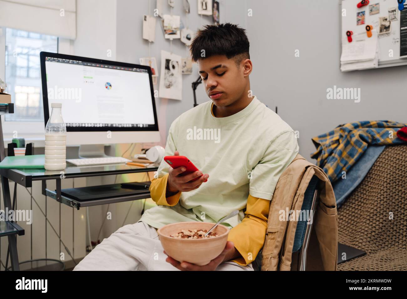 Brunette teenage boy using cellphone and eating breakfast in bedroom ...