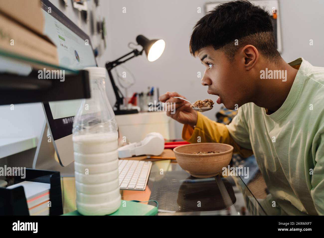 Brunette teenage boy using desktop computer while eating breakfast in ...