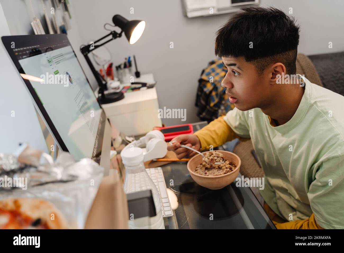Brunette teenage boy using desktop computer while eating breakfast in ...