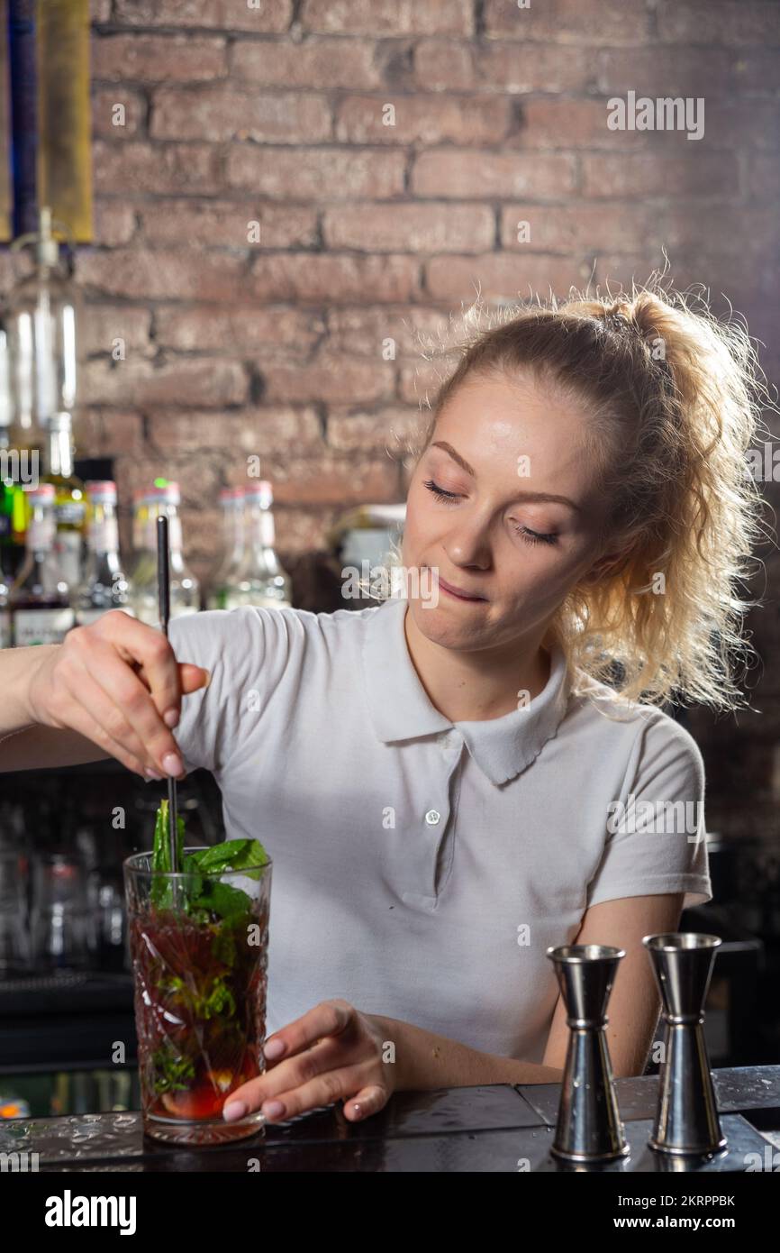 Young beautiful female bartender mixing a cocktail in a high glass ...