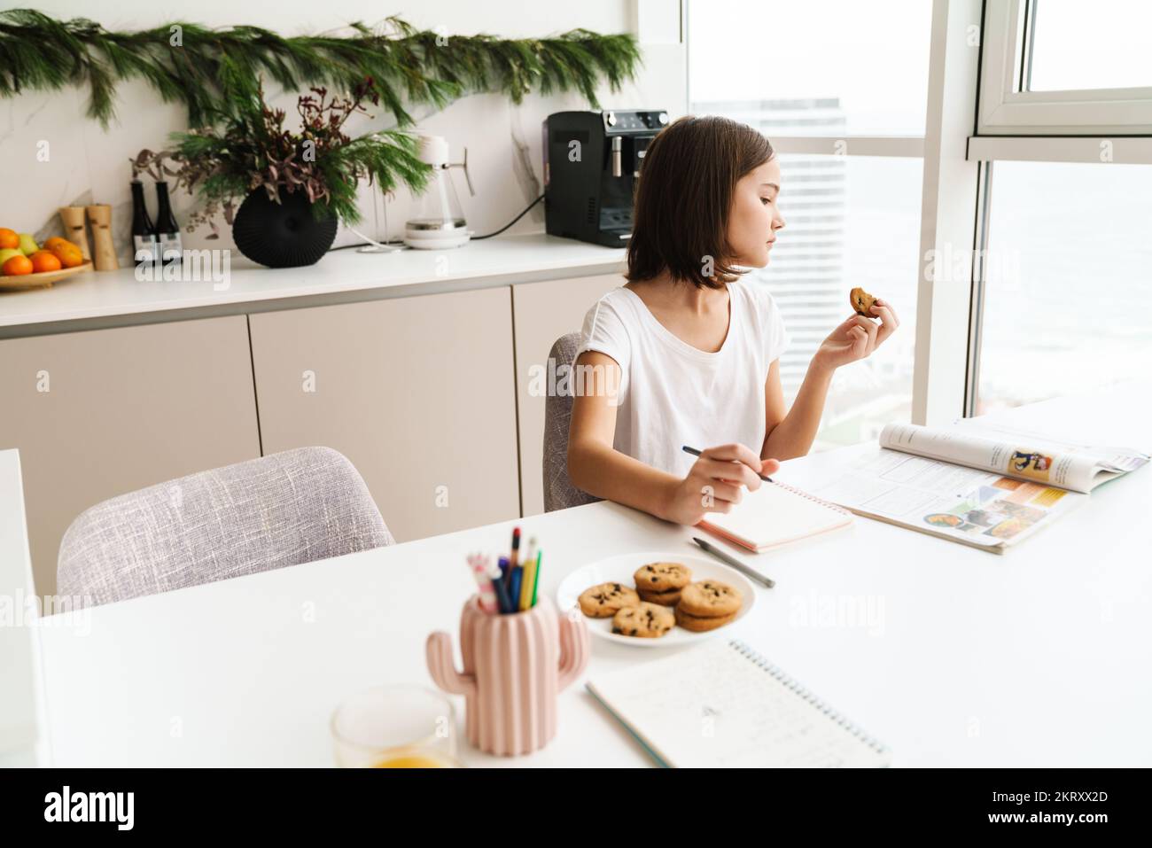 White preteen girl eating cookie while doing homework at home Stock ...