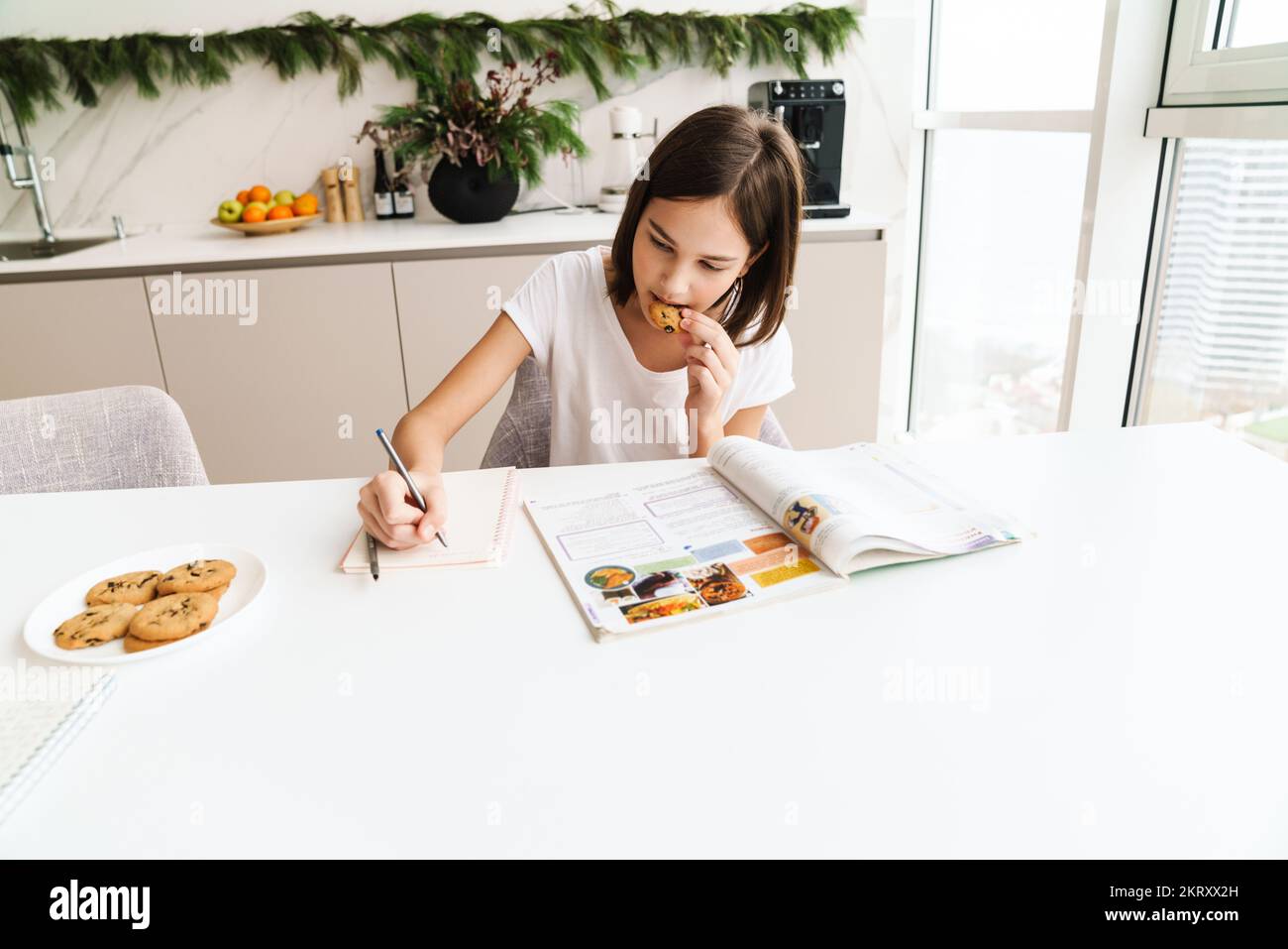 White preteen girl eating cookie while doing homework at home Stock ...