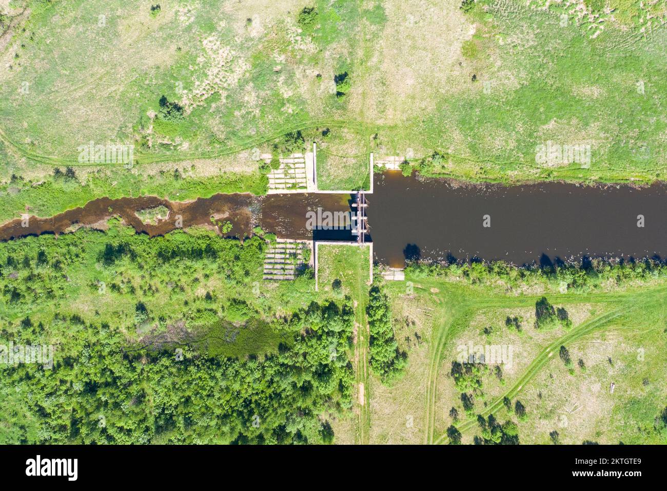 Small dam on a flat river flowing through green meadows, top aerial ...