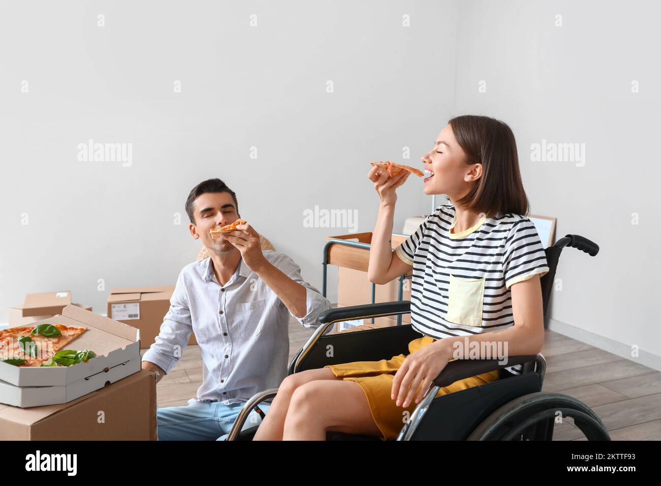Young man and his wife in wheelchair eating pizza in their new flat on ...