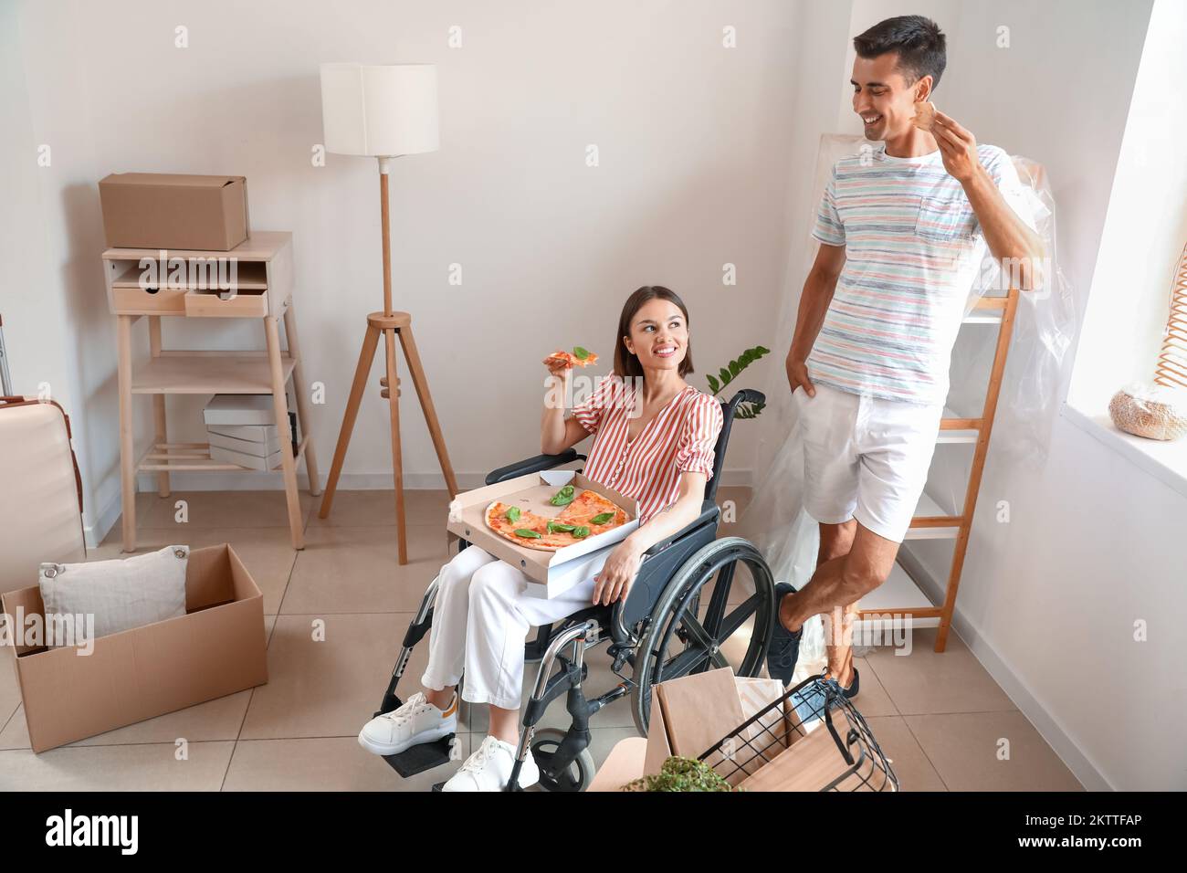 Young man and his wife in wheelchair eating pizza in room on moving day ...