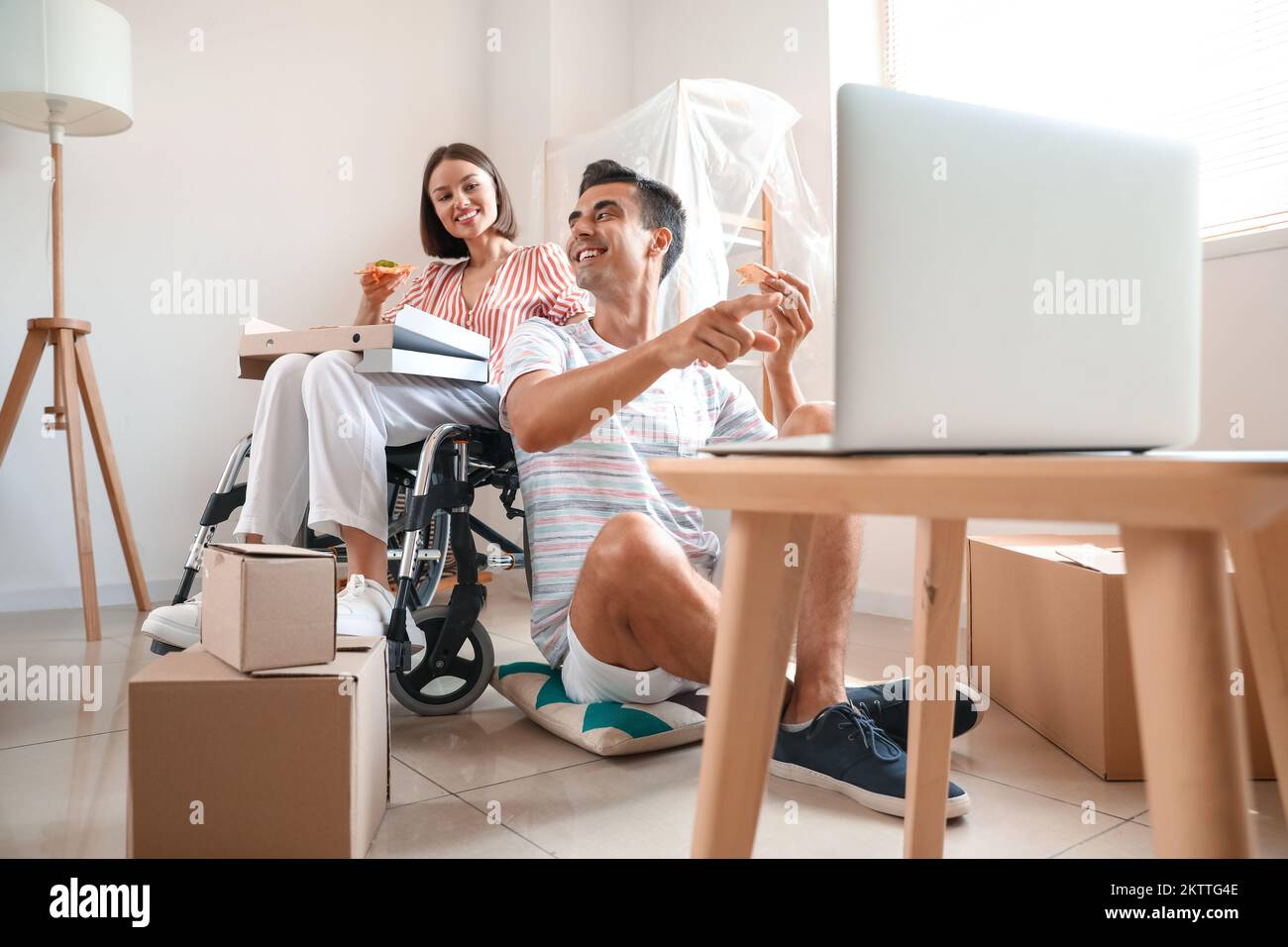 Young man and his wife in wheelchair with pizza watching video in room ...